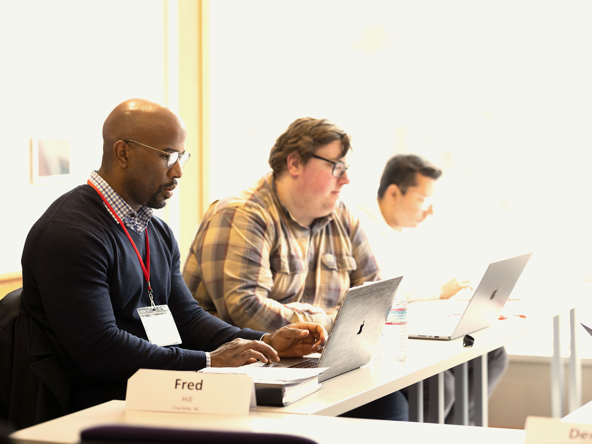 A man in a dark blue cardigan and checkered dress shirt sits at a classroom desk working on a laptop, with two other attendees beside him, all focused on their screens.